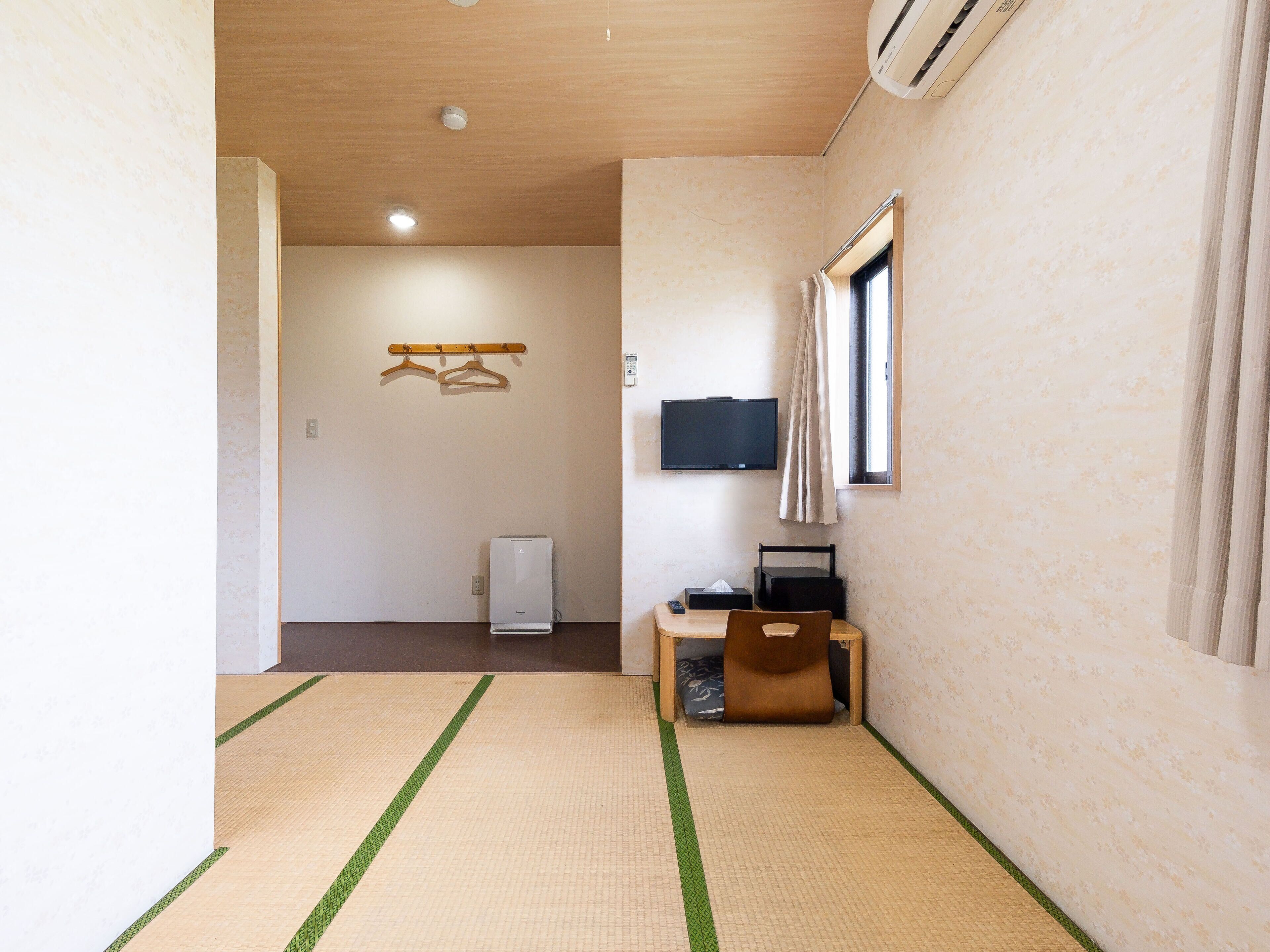 Quarto de hotel com piso de tatame, televisão na parede, uma pequena escrivaninha com cadeira e uma janela com cortinas.