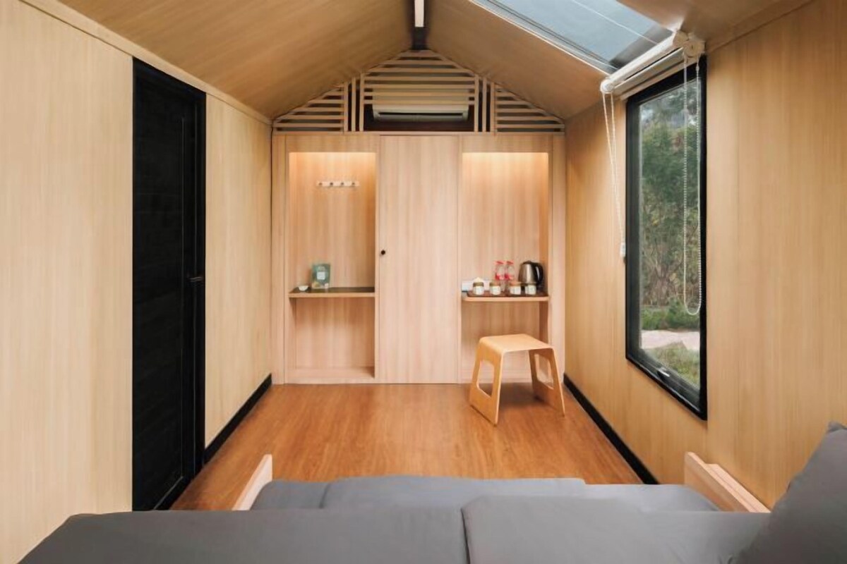 Quarto compacto e moderno, com paredes de madeira e teto inclinado, apresentando uma cama, um banquinho pequeno e uma janela com vista para o exterior.