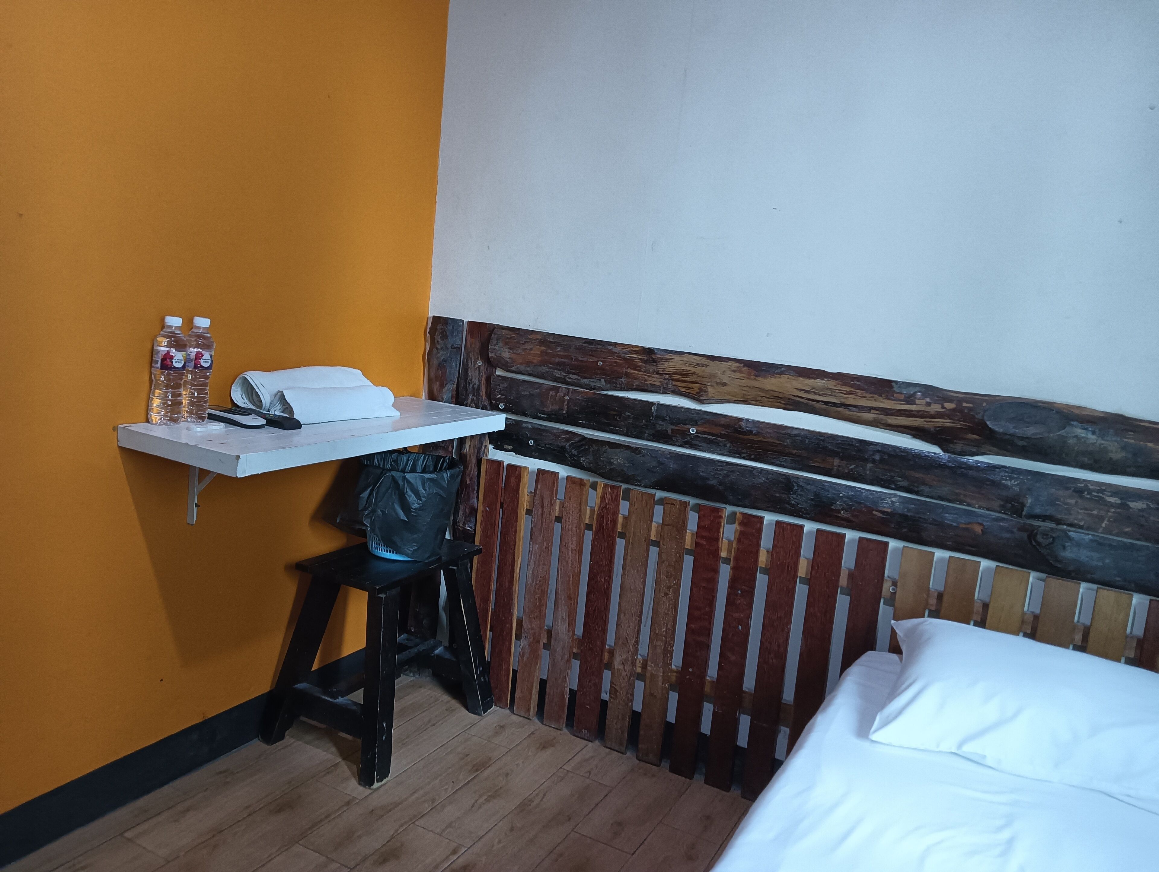 Um quarto pequeno com piso de madeira, uma cama branca, cabeceira de madeira, uma escrivaninha pequena com cadeira e uma prateleira com toalha e duas garrafas.