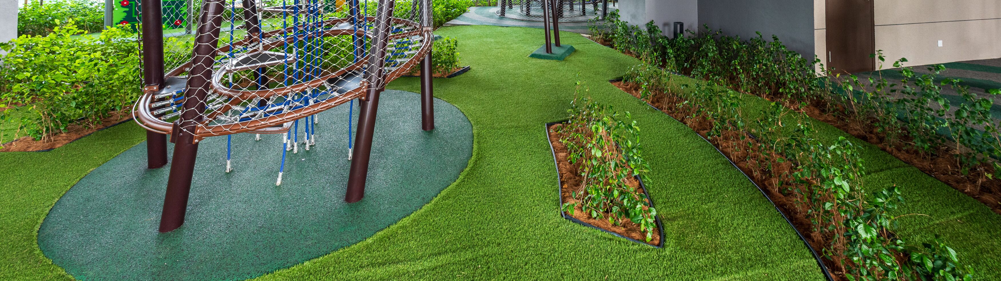 Um parquinho com uma ponte de corda e montes circulares de grama sintética, cercado por plantas verdes e um muro de concreto.