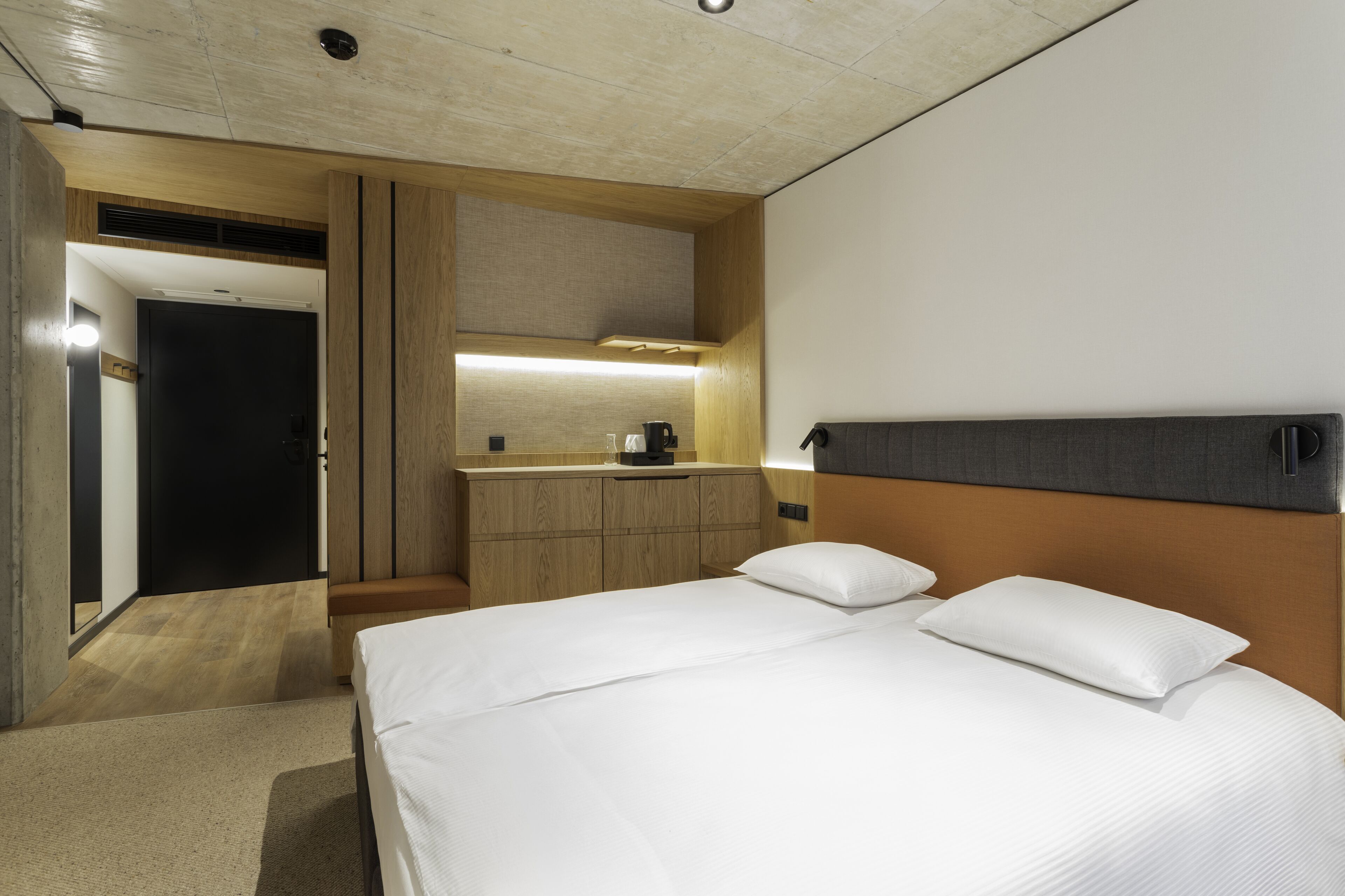 Quarto de hotel com cama, pequeno espaço de cozinha, porta e luminária no teto.