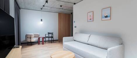 Uma sala de estar moderna com um sofá cinza, uma mesa de centro de madeira e uma TV sobre um suporte de madeira. Há duas cadeiras e uma mesa pequena ao fundo.