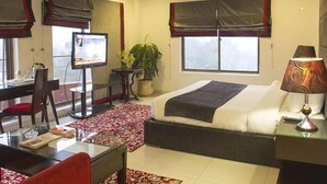 Quarto de hotel com cama, escrivaninha, cadeira, televisão e planta em vaso.