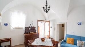 Uma sala de jantar com uma mesa de madeira, cadeiras e um candelabro. Na área de estar adjacente, há um sofá azul com almofadas.