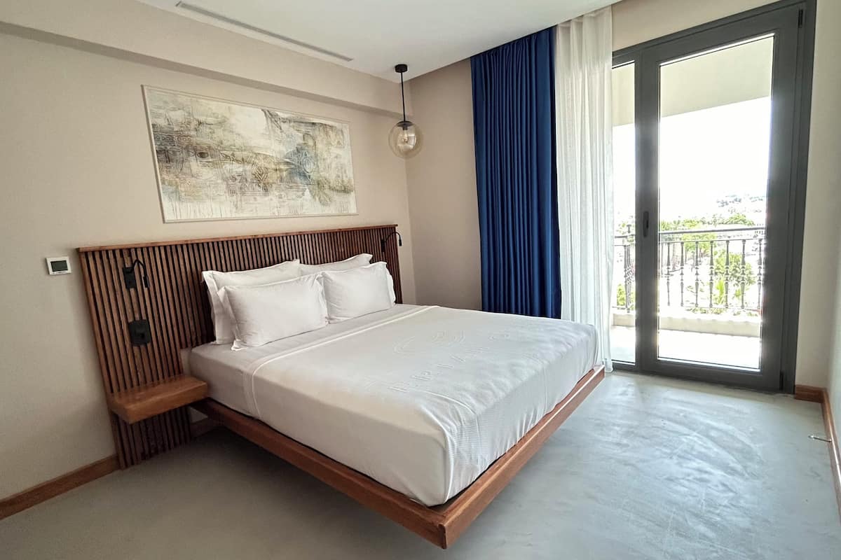 Quarto de hotel com estrutura de cama em madeira, roupa de cama branca, luminária no teto, uma janela grande com cortinas azuis e uma obra de arte pendurada na parede.