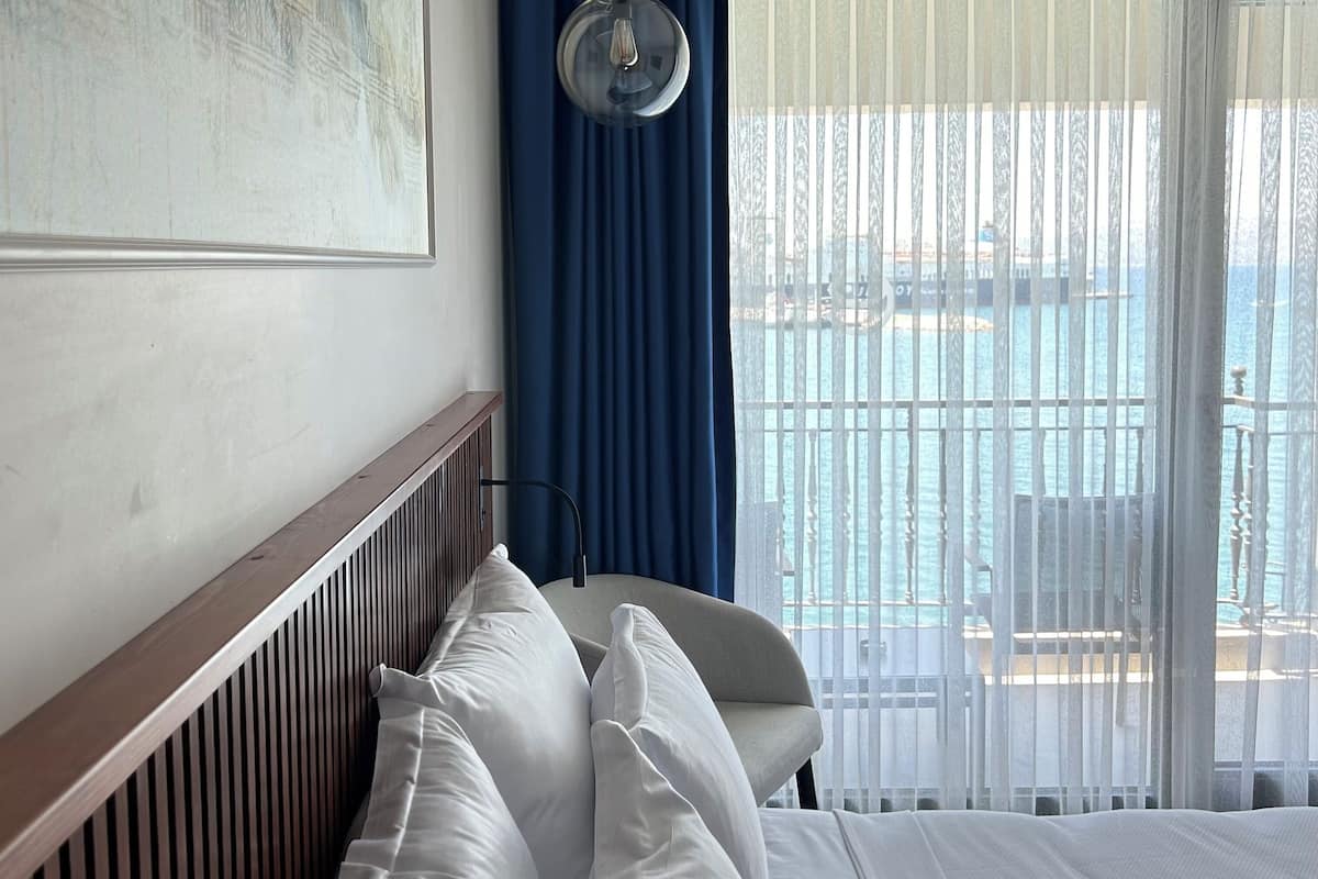 Quarto de hotel com cama, escrivaninha, cadeira, janela ampla com vista para uma piscina e varanda.