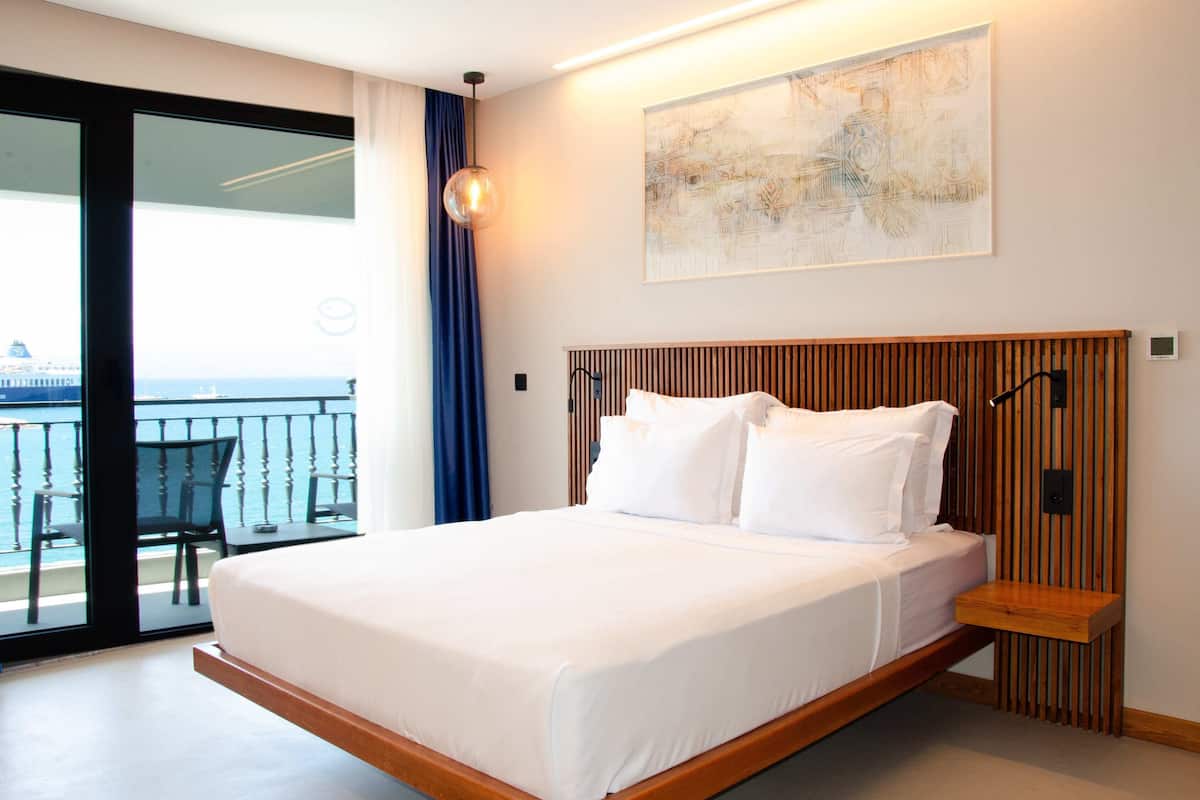 Quarto de hotel moderno com uma cama grande, cabeceira de madeira, varanda com vista para o mar e um quadro na parede.