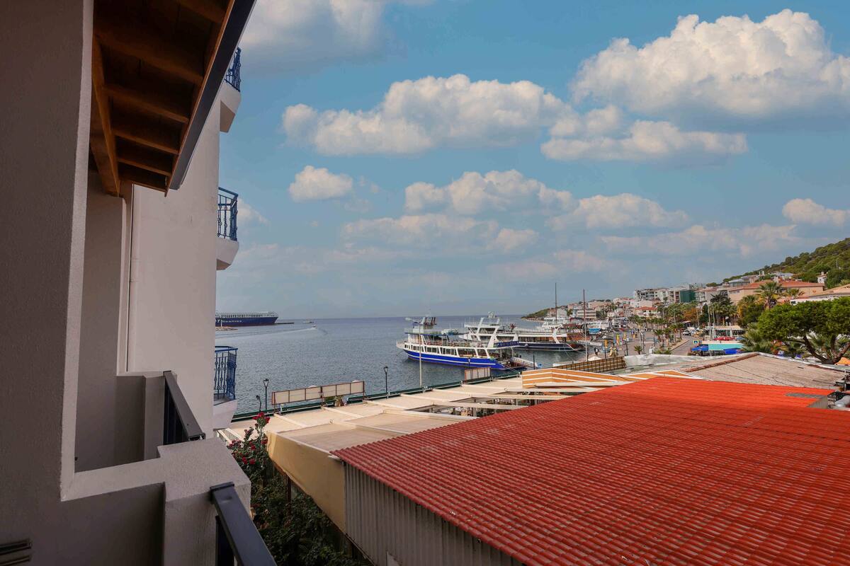 Vista costeira com um edifício de telhado vermelho, um cais com barcos e uma paisagem urbana ao fundo.