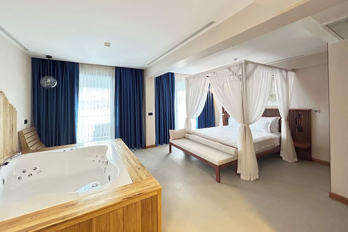 Quarto de hotel moderno com uma banheira grande, uma cama de casal com dossel e um sofá separado.