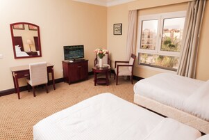 Quarto de hotel com cama, escrivaninha e cadeira, televisão, pequena mesa com um vaso de flores e janela com vista para prédios.