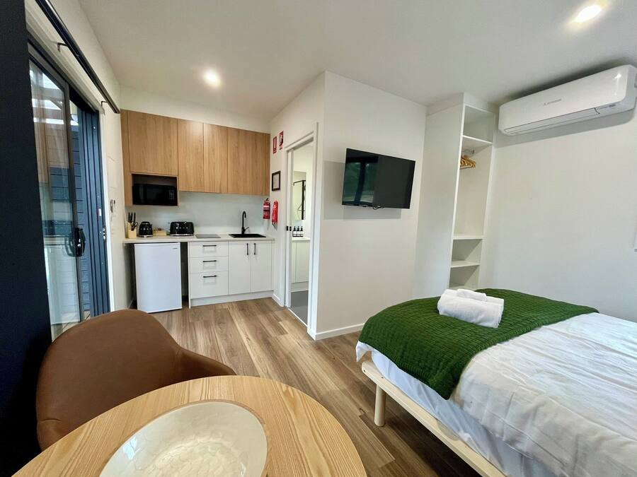 Quarto de hotel compacto com cozinha compacta, uma cama com manta verde, TV fixada na parede e uma pequena área de refeições.