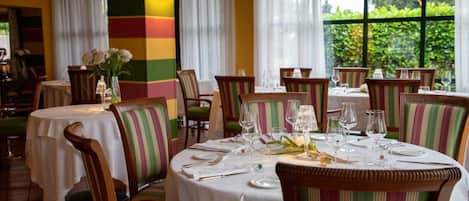 Restaurante com mesas redondas preparadas para refeições, cadeiras listradas e vista para o verde através de grandes janelas.
