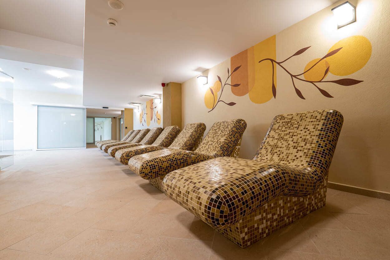 Fileira de cadeiras de descanso com padrão de mosaico em um corredor amplo e bem iluminado, com design moderno e um mural na parede.
