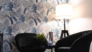 Um quarto com papel de parede estampado, uma poltrona preta, uma mesinha com uma planta e um castiçal, e um abajur.
