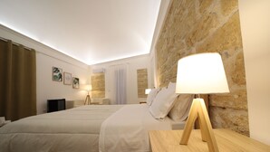 Quarto de hotel com uma cama grande, mesa de cabeceira com abajur, televisão e parede de pedra.