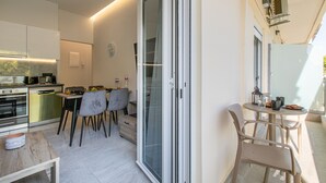 Cozinha moderna com eletrodomésticos de aço inoxidável, área de jantar com mesa e cadeiras, e porta de vidro deslizante que dá para uma varanda.