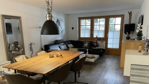Uma área de refeições com mesa e cadeiras de madeira, uma luminária pendente preta e um sofá com almofadas ao fundo.
