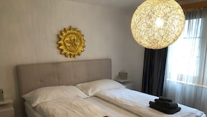 Uma cama de casal com roupa de cama branca e duas toalhas dobradas, um ornamento dourado em formato de sol na parede, e um candeeiro suspenso.