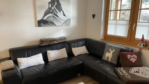 Um sofá de couro preto com almofadas estampadas e lisas, um tapete branco e uma obra de arte na parede retratando uma montanha.