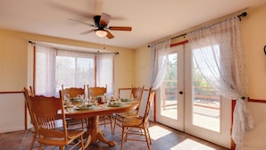 Uma sala de jantar com mesa e cadeiras de madeira, ventilador de teto e grandes janelas com cortinas.