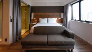 Quarto de hotel moderno com uma cama grande, um sofá, área de chuveiro em vidro e vista para a cidade.