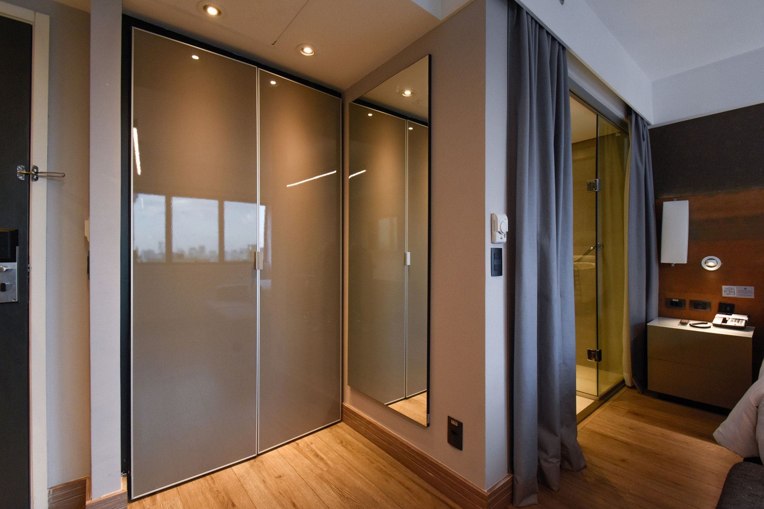Quarto de hotel moderno com closet de porta deslizante, piso de madeira, mesa de cabeceira pequena com luminária, e vista para a cidade pela janela.
