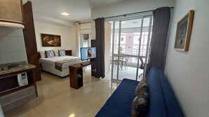 Quarto de hotel com cama, televisão, sofá, varanda com vista para edifícios e área de refeições.