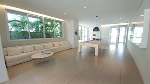 Uma sala de estar moderna com um sofá bege, uma mesa de centro redonda e uma mesa de bilhar. Grandes janelas proporcionam luz natural e vista para o verde lá fora.