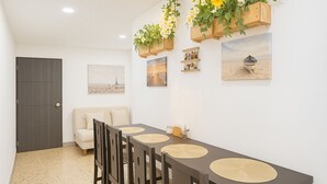 Uma área de refeições com uma mesa comprida, cadeiras e um sofá. Há prateleiras de madeira com plantas e quadros na parede.