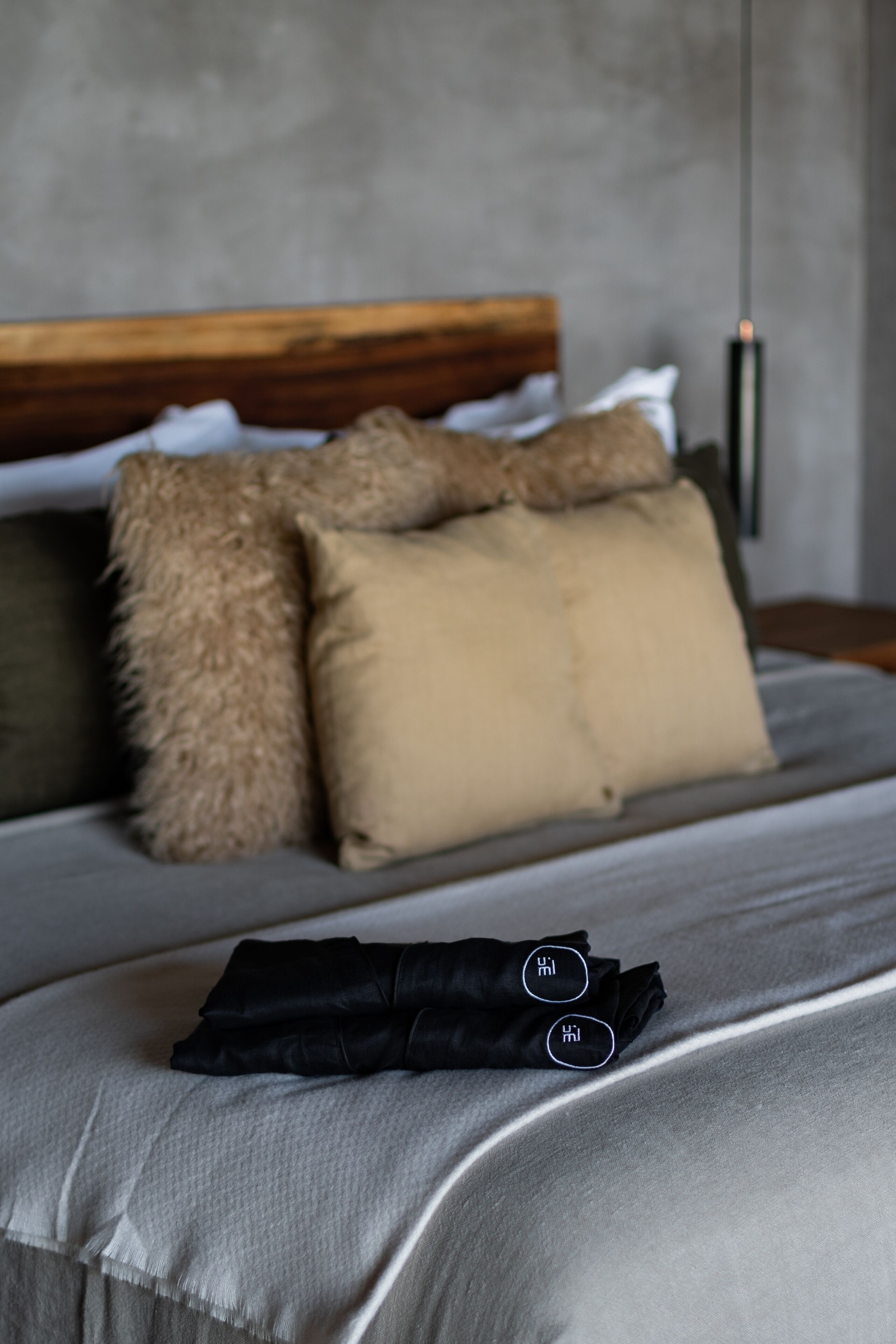 Cama bem arrumada com colcha cinza, dois travesseiros e uma peça de roupa preta dobrada sobre ela.
