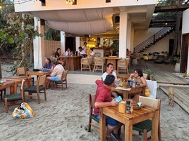 Uma área externa com mesas e cadeiras de madeira, uma seção coberta e um bar com pessoas.