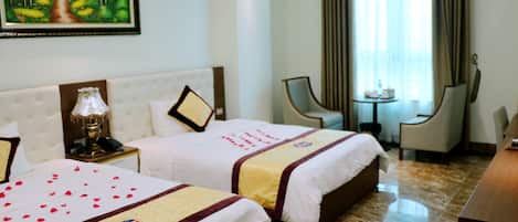 Quarto de hotel com duas camas, uma mesa de cabeceira com abajur, uma pequena mesa, duas cadeiras e um quadro na parede.