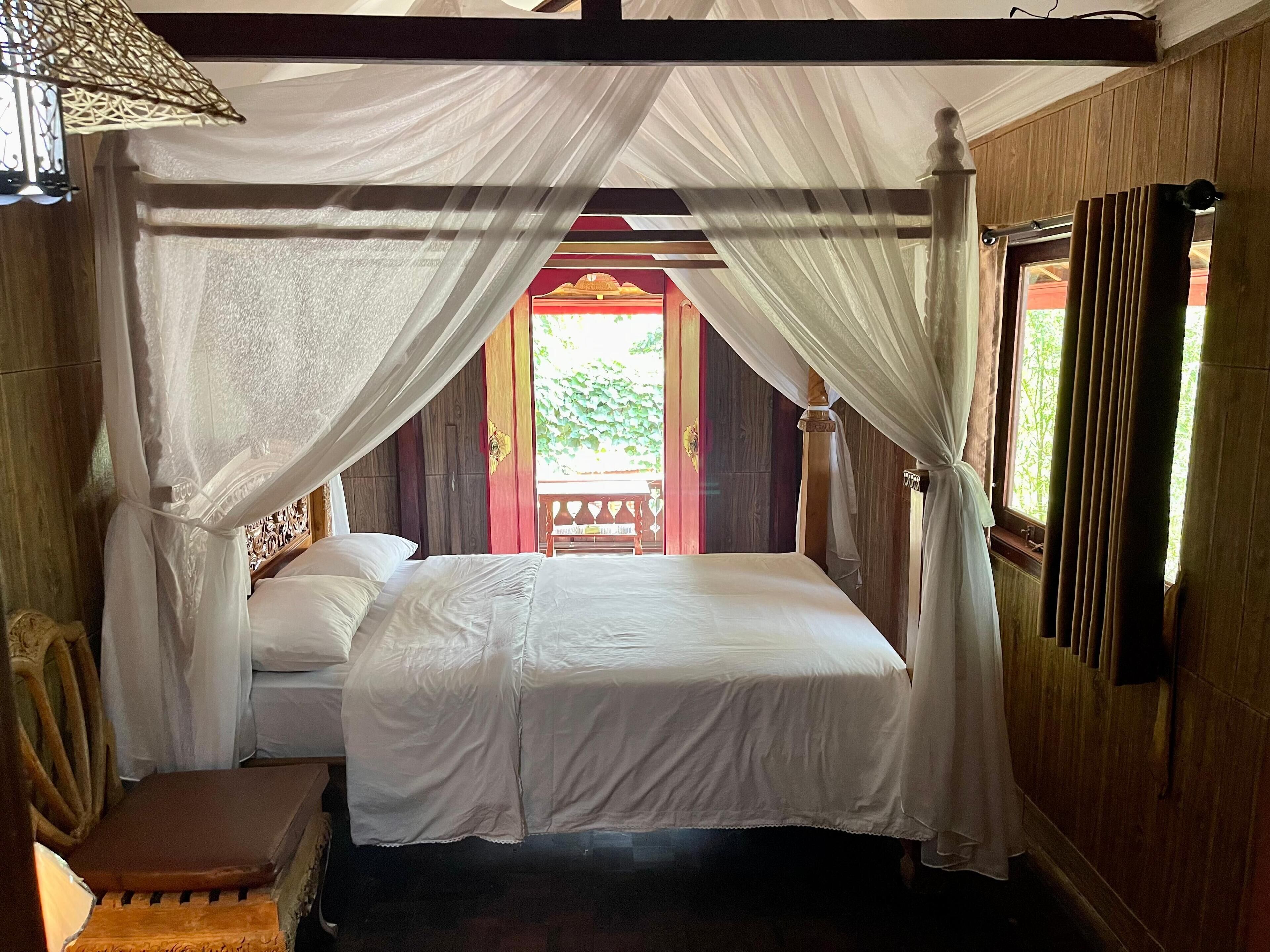 Quarto com cama sob um dossel, paredes de madeira e vista para um edifício vermelho lá fora.