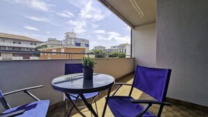 Uma varanda com uma mesa redonda e duas cadeiras, um vaso com planta e vista para edifícios ao fundo.