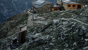 Uma pousada de montanha com vista desobstruída para picos nevados, um pequeno edifício com telhado inclinado e um mastro.