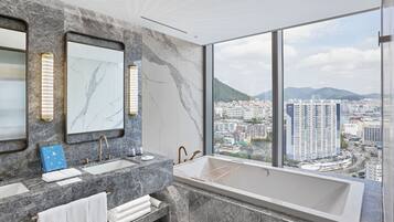 Casa de banho moderna com uma banheira grande, duas pias e um espelho amplo. Apresenta paredes de mármore e vista para a cidade através de grandes janelas.