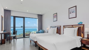 Quarto de hotel com duas camas, uma escrivaninha e uma cadeira. Há uma janela grande com vista para o oceano.