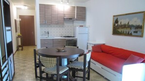 Cozinha compacta com eletrodomésticos modernos, área de refeições com mesa redonda e cadeiras, e um sofá vermelho encostado na parede.