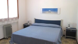 Quarto com uma cama, duas mesas de cabeceira, um radiador, uma janela com cortinas de renda e um quadro na parede.