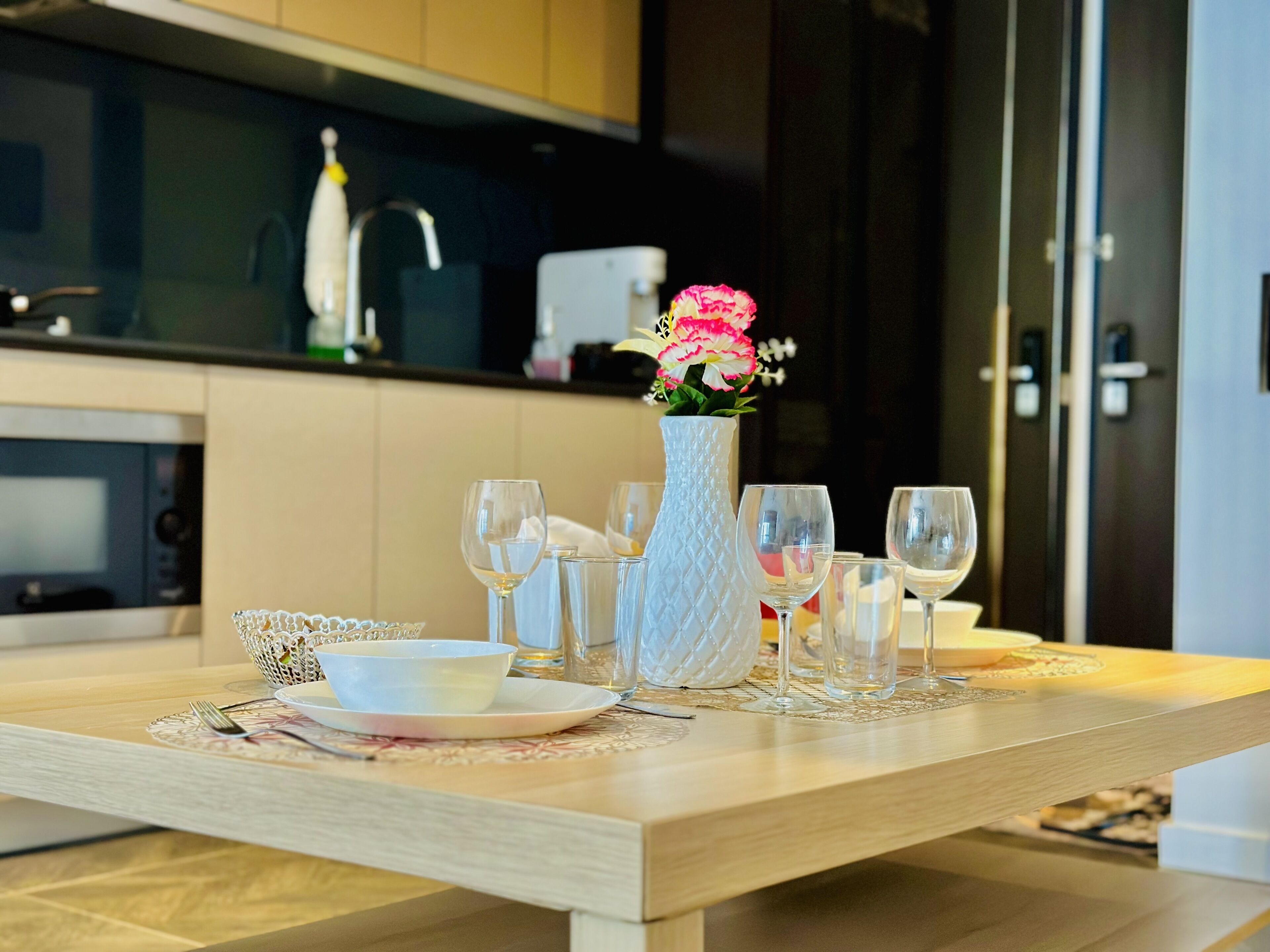 Mesa de jantar posta com pratos, tigelas e copos de vinho brancos, um vaso branco com flores cor-de-rosa e uma cesta decorativa.