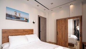 Quarto de hotel com cabeceira de madeira, uma cama grande com roupa de cama branca, uma luminária na parede, uma porta com espelho e um quadro com uma imagem de um cais.