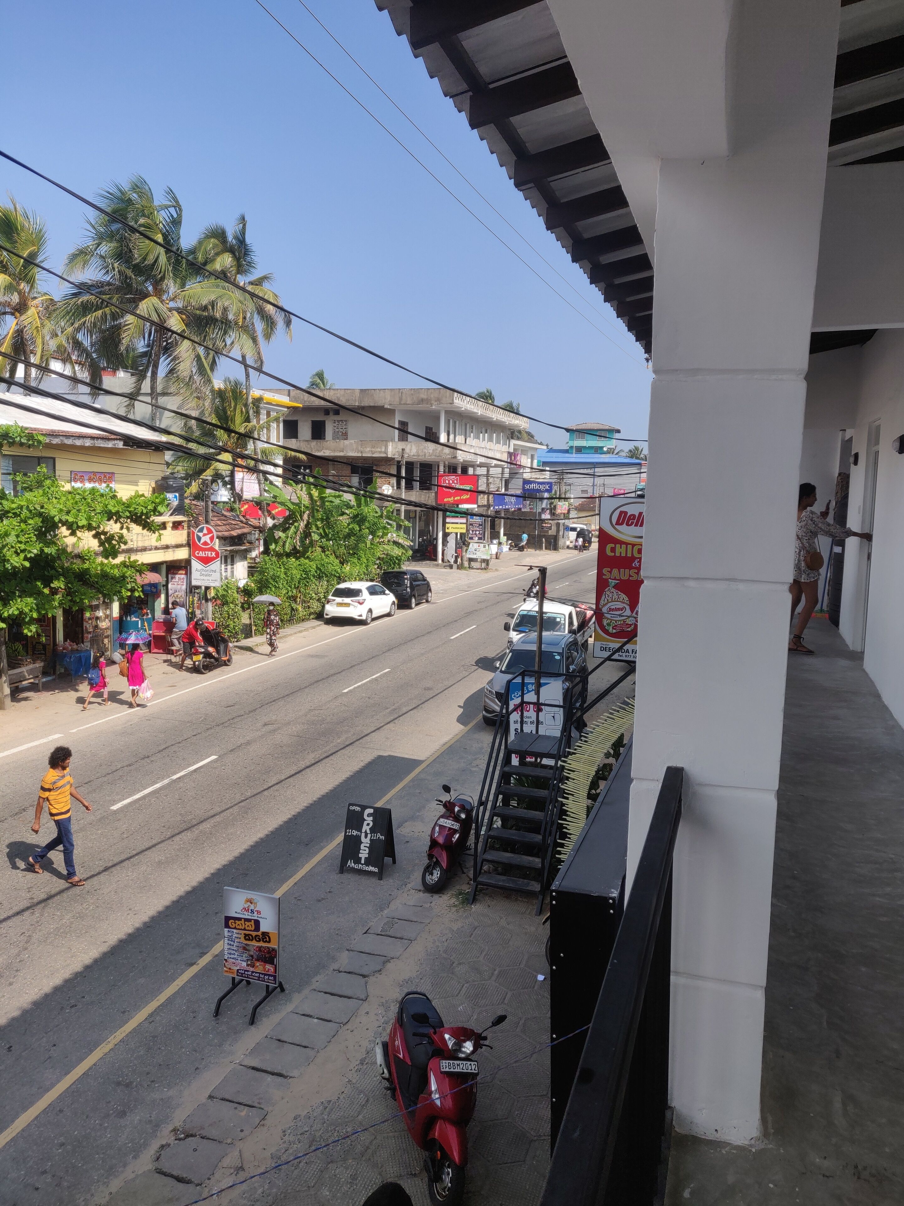 Vista da rua com uma scooter vermelha estacionada na calçada, uma faixa de pedestres e diversos estabelecimentos comerciais e edifícios ao fundo.