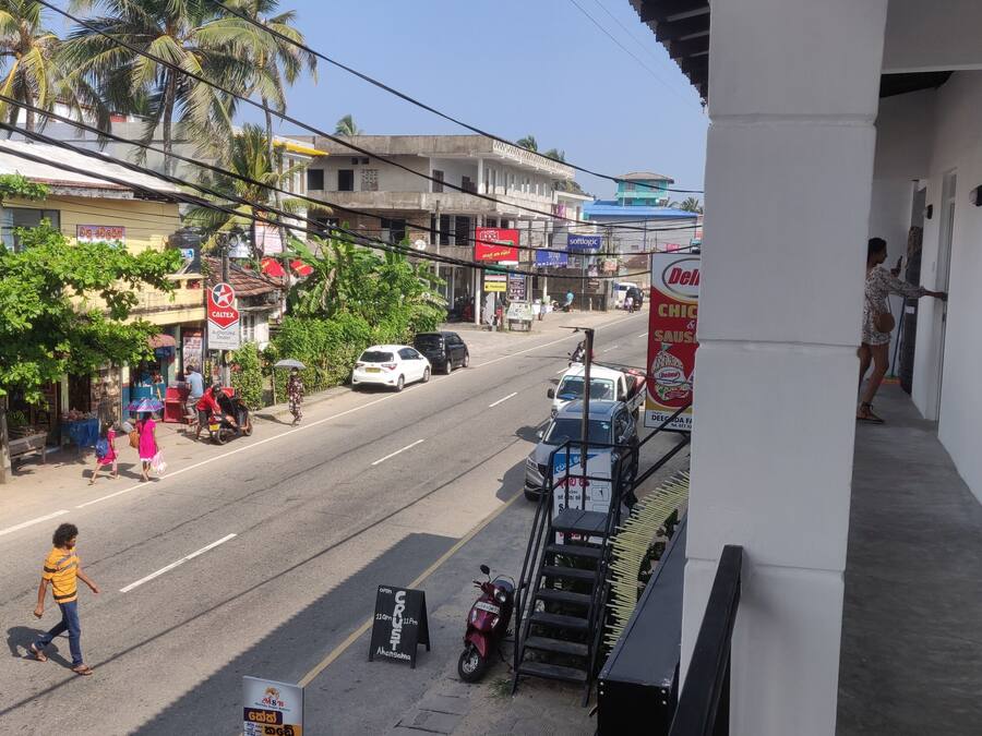 Vista da rua com uma scooter vermelha estacionada na calçada, uma faixa de pedestres e diversos estabelecimentos comerciais e edifícios ao fundo.