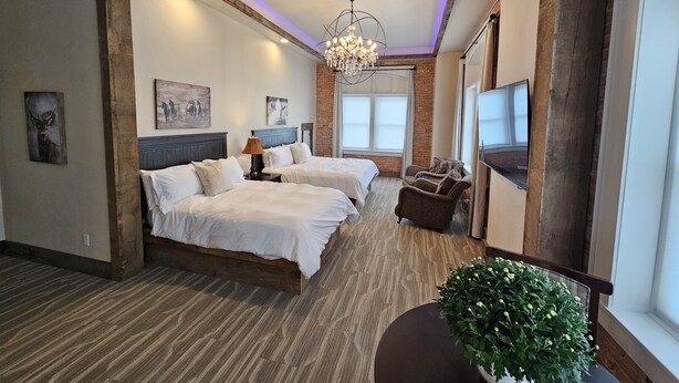 Quarto de hotel moderno com uma cama grande, área de estar, lustre e televisão fixada na parede.