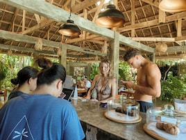 Restaurante com vigas de madeira e telhado de palha, com balcão e cúpula de vidro para exposição de alimentos, e uma pessoa servindo.