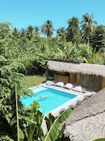 Piscina com espreguiçadeiras e almofadas, rodeada por vegetação exuberante e palmeiras.