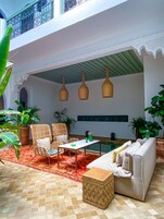 Amplo espaço ao ar livre com um sofá branco, cadeiras de vime e uma mesa. Há luminárias pendentes, plantas em vasos e piso de azulejo.
