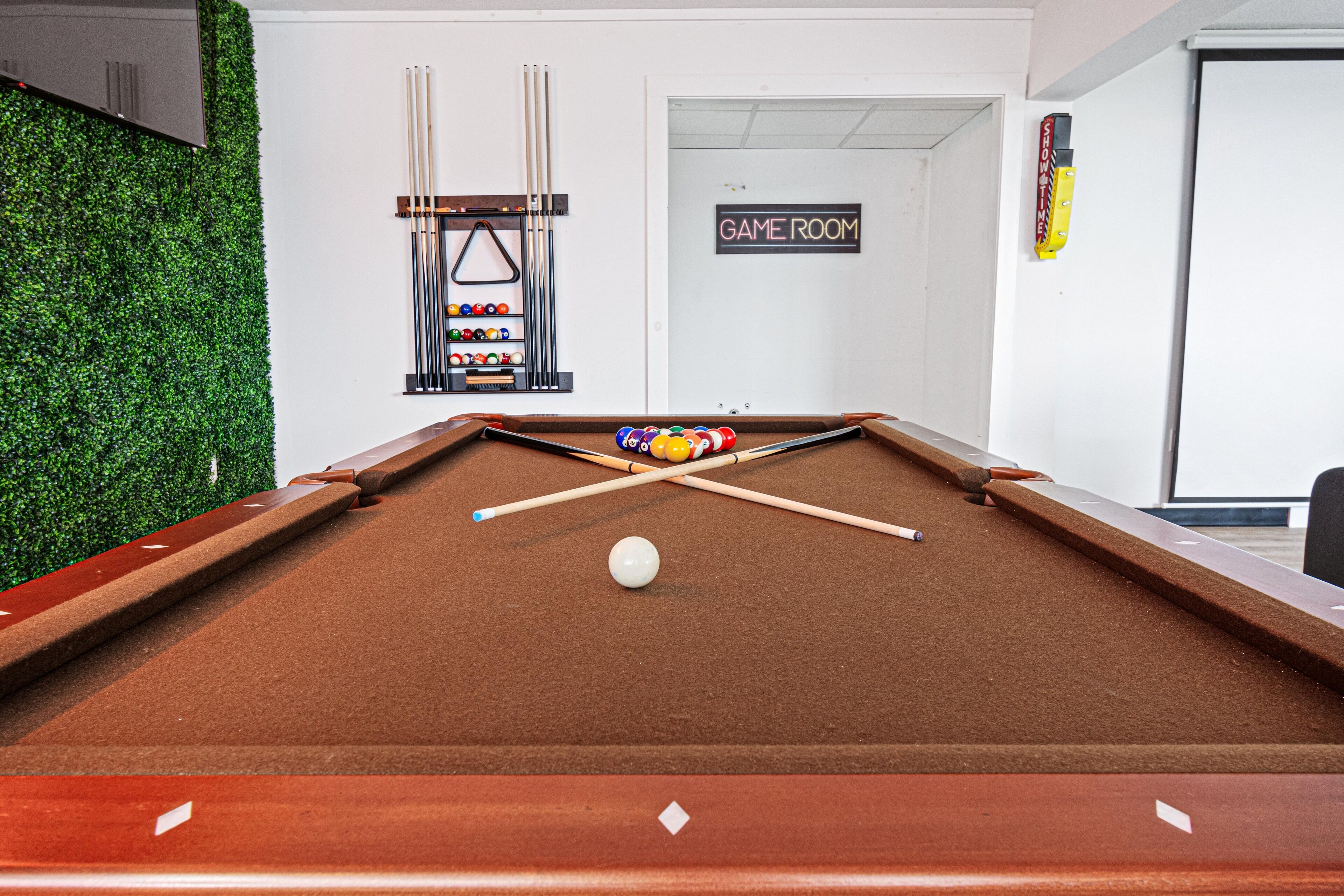 Uma mesa de bilhar com bolas e tacos, uma parede verde e um aviso que diz 'GAME ROOM'.