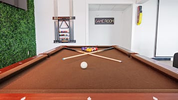 Uma mesa de bilhar com bolas e tacos, uma parede verde e um aviso que diz 'GAME ROOM'.