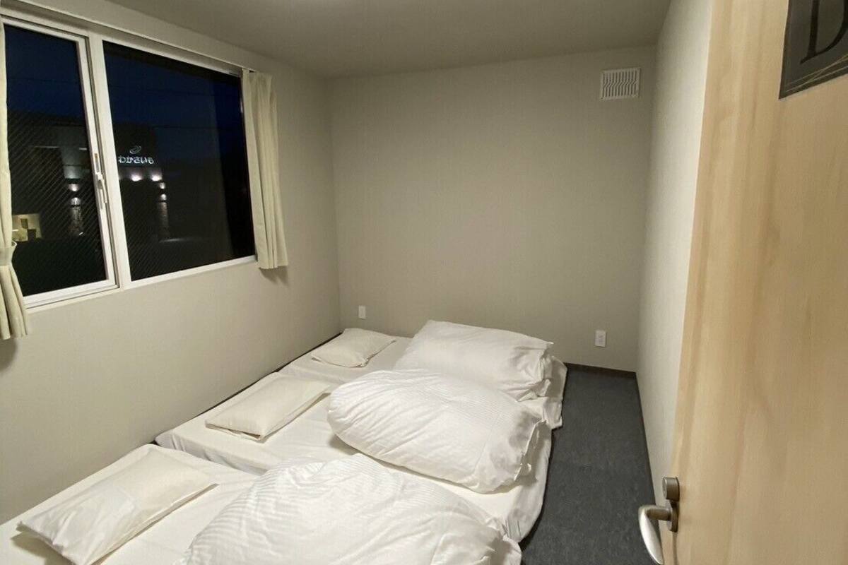 Uma cama de solteiro com roupa de cama branca num quarto com janela, porta com placa 'B' e luz no teto.